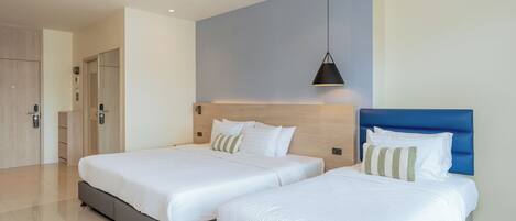 Quarto de hotel com duas camas, uma cadeira azul e uma luminária pendente moderna.