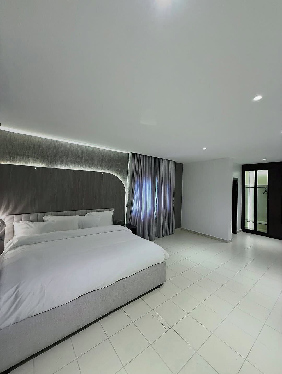 Quarto moderno com cama grande, mesa de cabeceira, janela com cortinas e porta para outro cômodo.