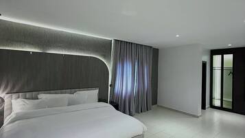 Quarto moderno com cama grande, mesa de cabeceira, janela com cortinas e porta para outro cômodo.