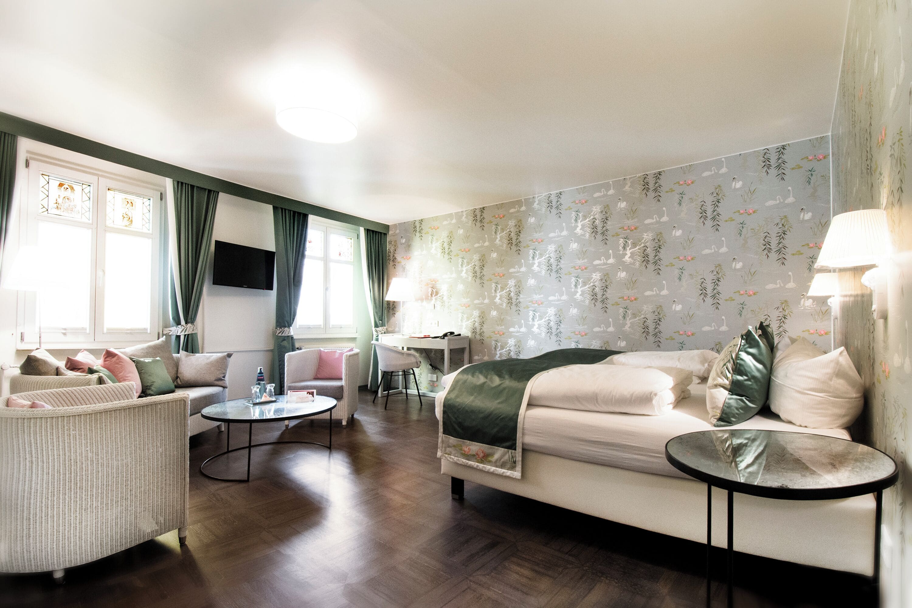 Uma sala de estar acolhedora com sofá, poltrona e mesa de centro. Há uma escrivaninha com cadeira e luminária. O quarto tem uma janela com cortinas e uma parede com padrão floral.