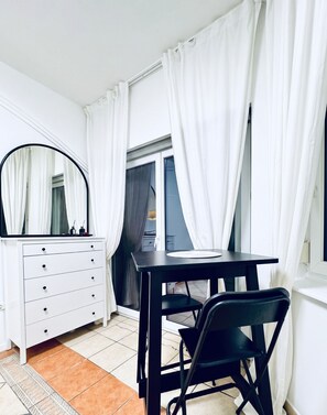 Um quarto com cômoda branca, espelho redondo, escrivaninha preta, cadeira preta e cortinas brancas.