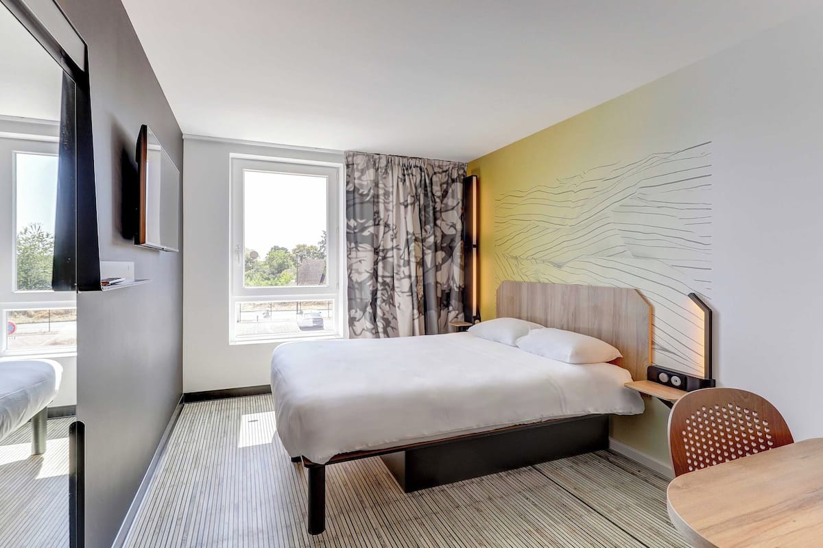 Quarto de hotel moderno com uma cama grande, uma mesa com cadeira, uma televisão de tela plana e uma janela com cortinas.