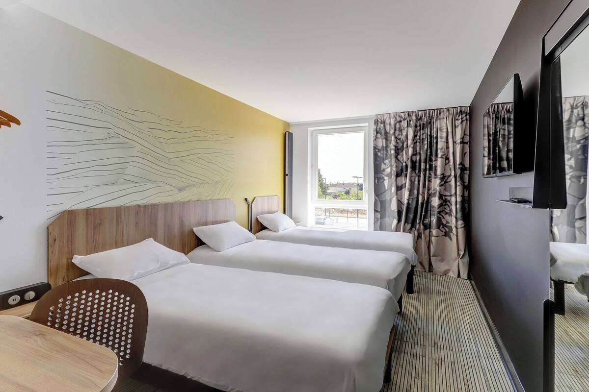 Quarto de hotel com duas camas, cabeceira de madeira, uma obra de arte na parede, janela com cortinas e televisão montada.