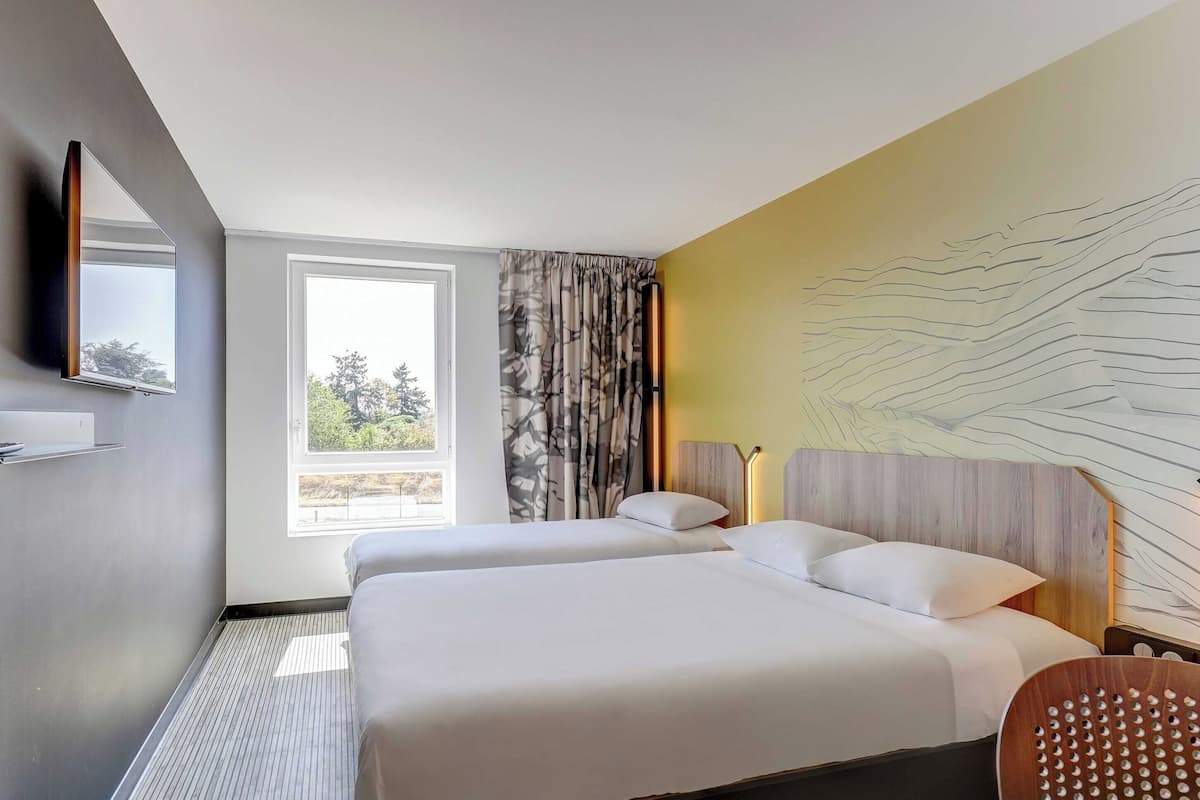 Quarto de hotel com duas camas, uma janela grande, televisão fixada na parede e uma obra de arte original.
