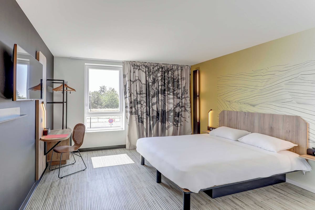 Quarto de hotel com cama, escrivaninha e cadeira, janela com cortinas e TV fixada na parede.