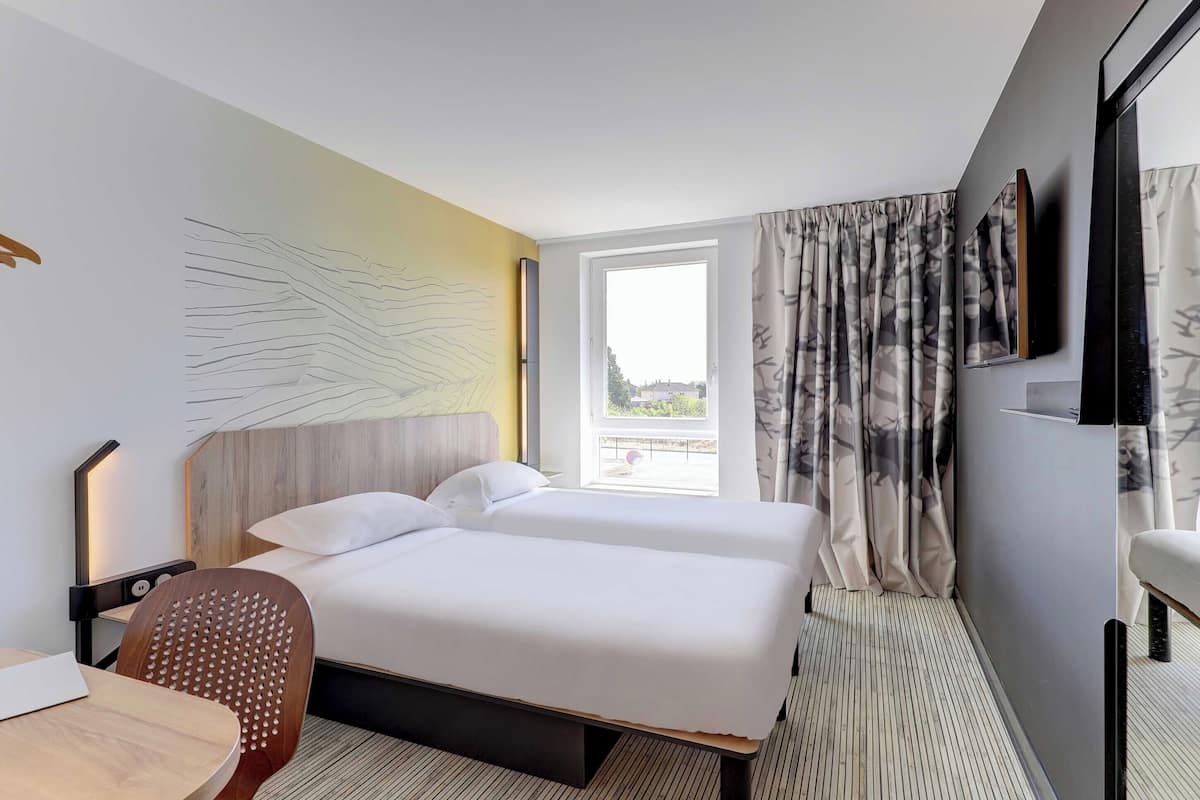 Quarto de hotel com cama, escrivaninha, cadeira, janela com cortinas e uma obra de arte na parede.