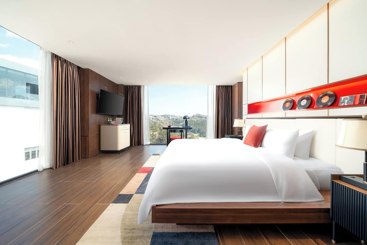 Quarto de hotel moderno com uma cama grande, televisão de tela plana, mesa de cabeceira e vista para a cidade.
