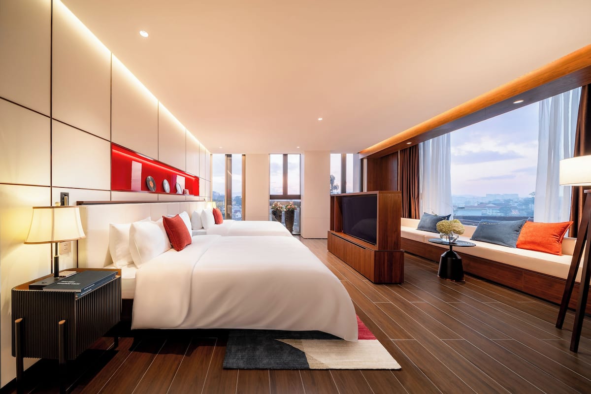 Quarto de hotel moderno com uma cama grande, televisão, área de estar com vista e uma parede de destaque vermelha.