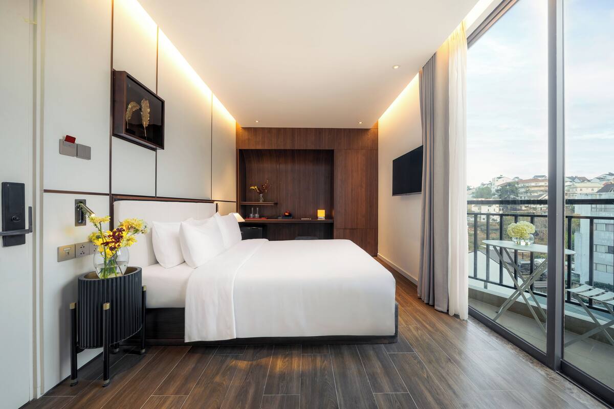 Quarto de hotel moderno com uma cama grande, uma pequena mesa de escritório, uma janela ampla com varanda e vista para a cidade.
