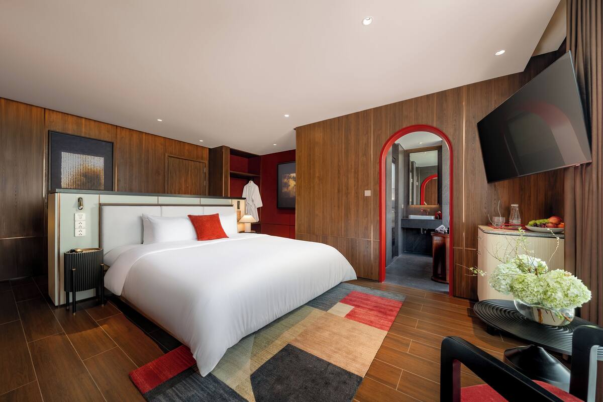 Quarto de hotel moderno com uma cama grande, uma escrivaninha com cadeira, televisão na parede e uma pequena mesa com um vaso de flores.