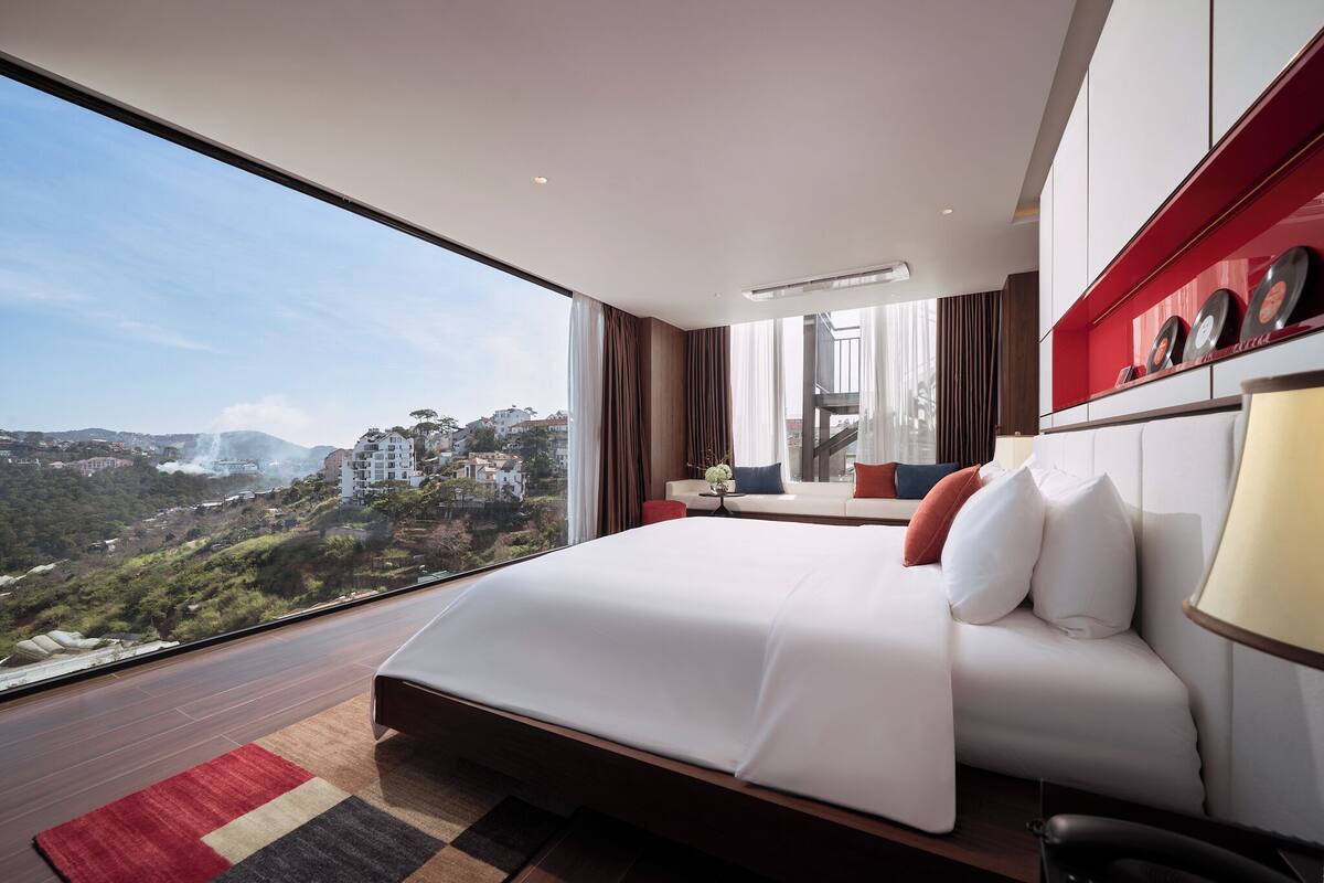 Quarto de hotel moderno com cama grande, vista para a cidade e uma parede de destaque vermelha.