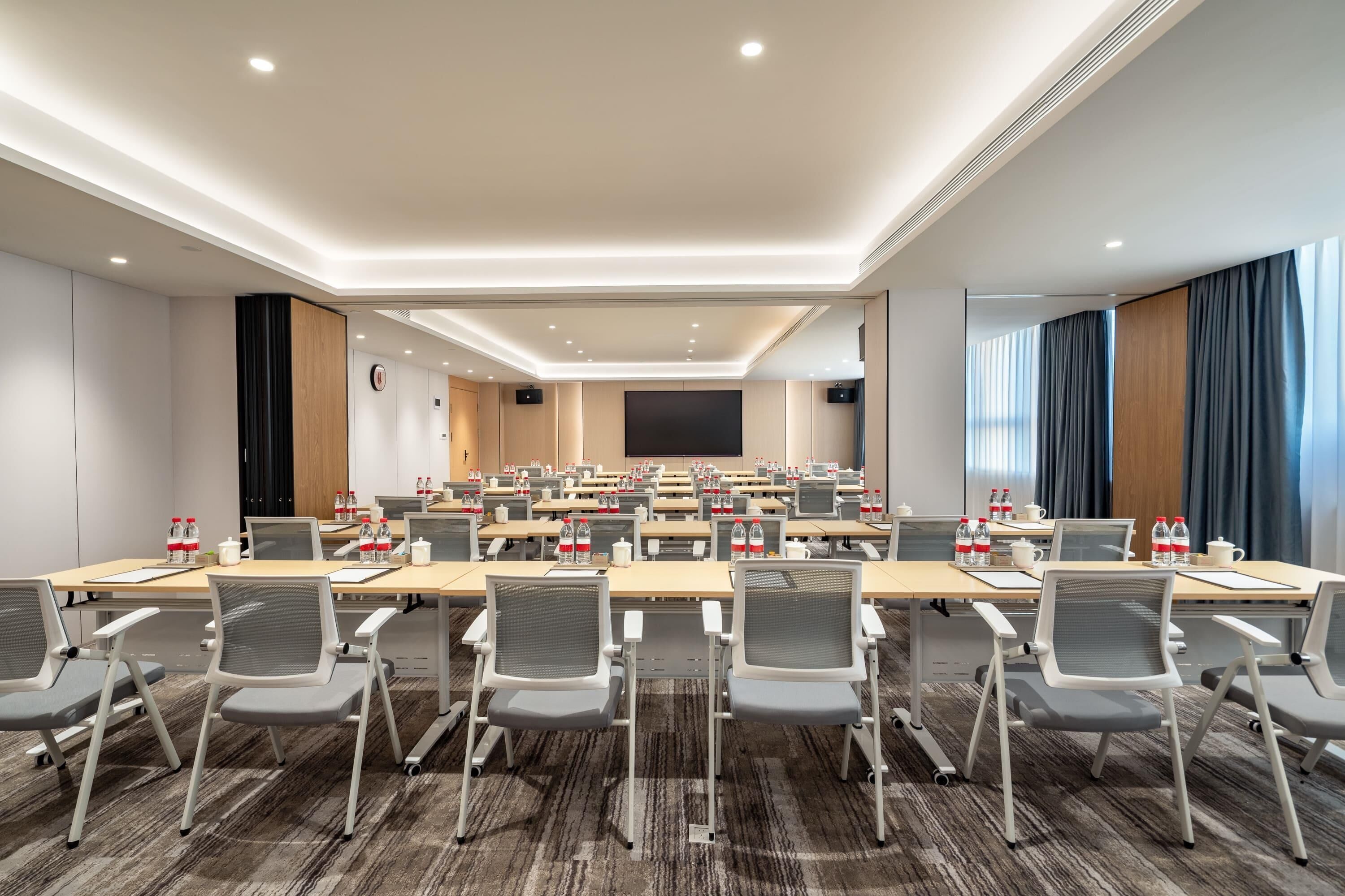 Sala de conferência com várias mesas e cadeiras, equipada com garrafas de água e bules, e uma tela grande na frente.