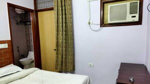 Quarto com cama, penteadeira de madeira, janela com ar condicionado, porta e banheiro visível por uma porta aberta.