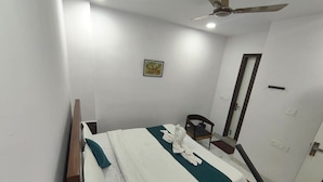 Quarto de hotel com uma cama de solteiro, um ventilador de teto, uma pequena escrivaninha com cadeira e uma porta.