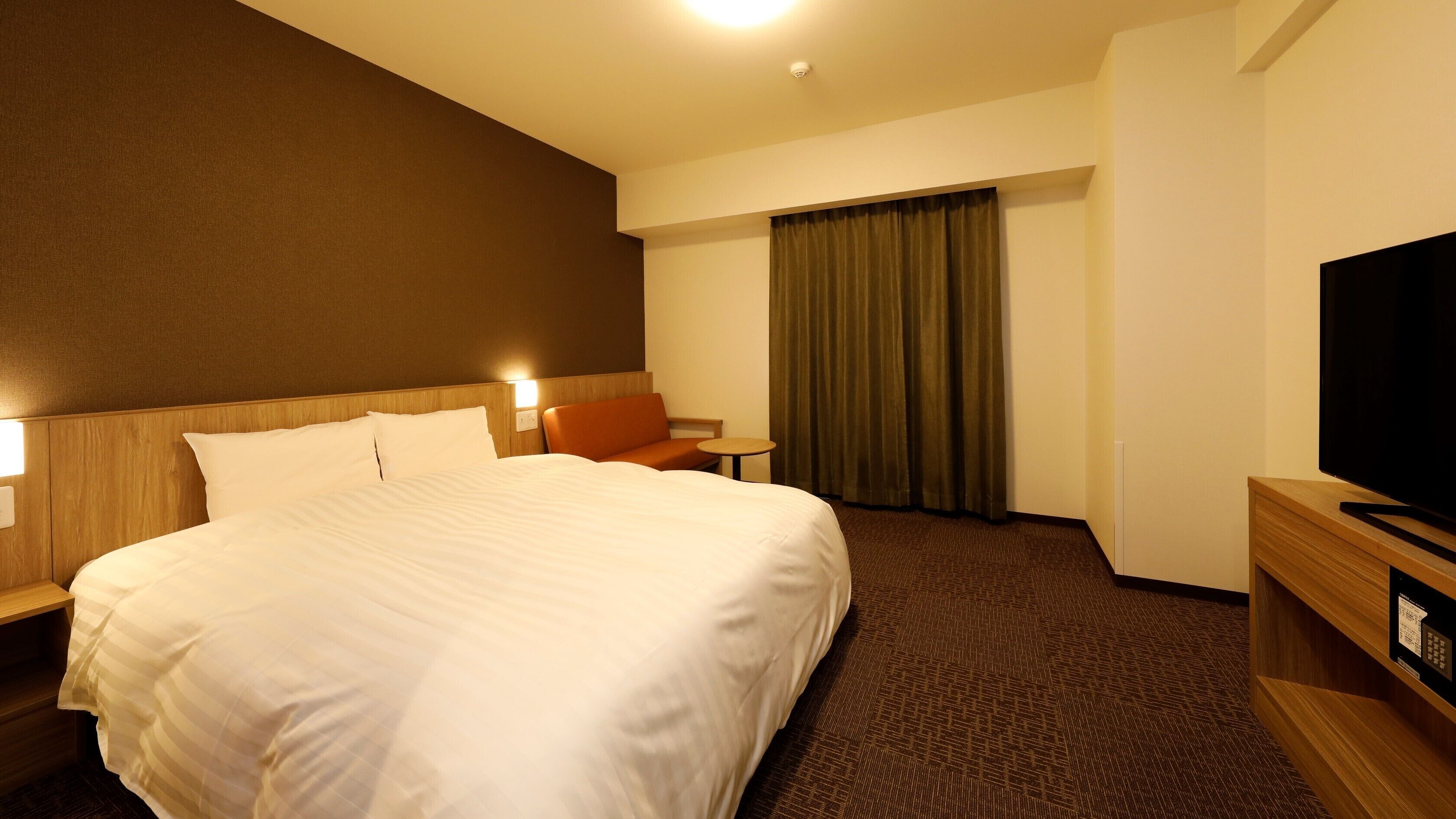 Quarto de hotel com uma cama grande, televisão, escrivaninha, cadeira e janela com cortinas.