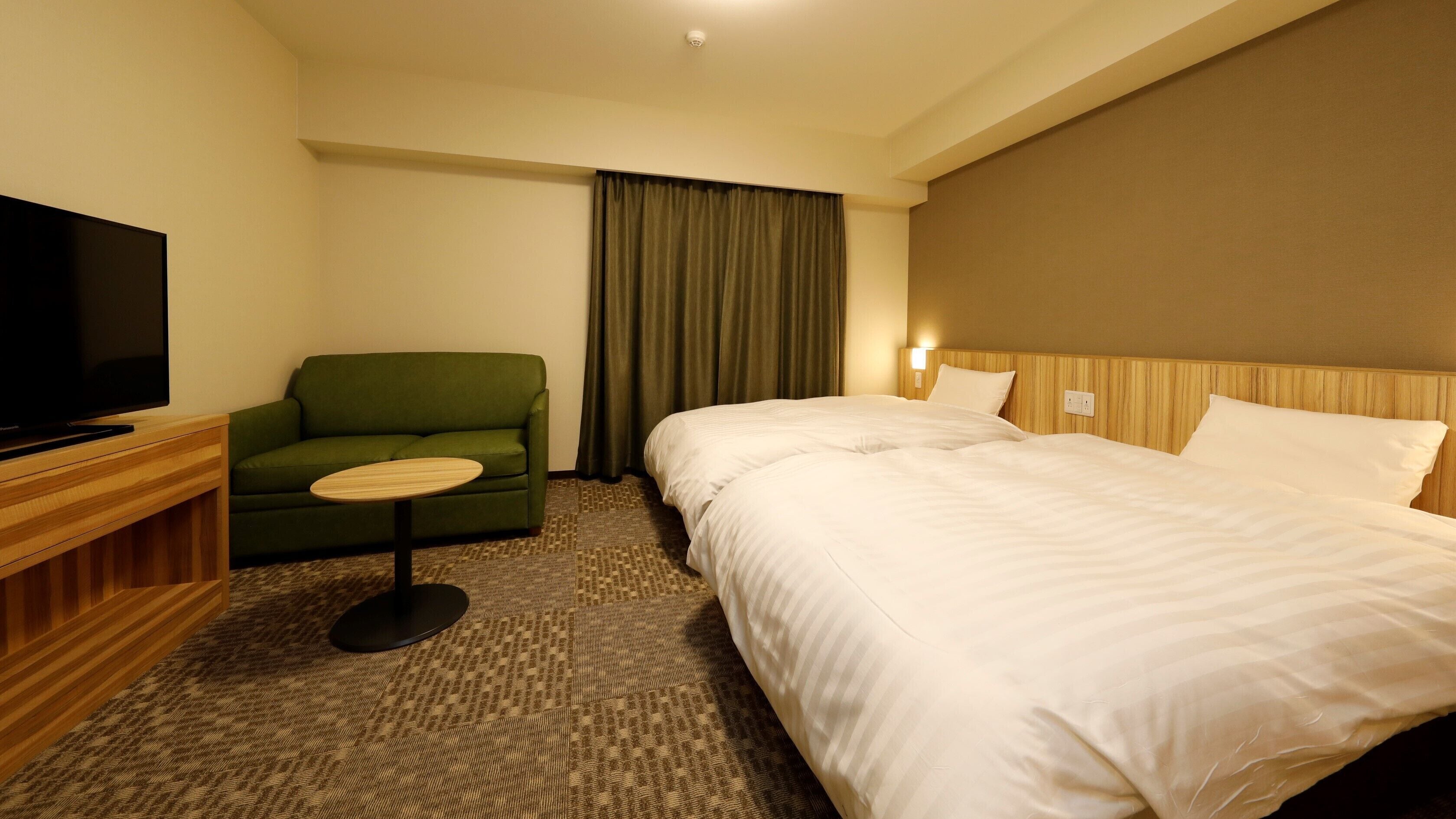 Quarto de hotel com uma cama, uma poltrona verde, uma mesa redonda pequena, uma televisão e uma janela com cortinas.