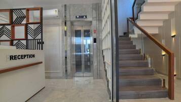 Uma área de recepção com balcão de atendimento identificado como "RECEPTION", uma escada com corrimãos de madeira e um elevador com parede espelhada.