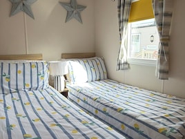 Quarto com duas camas, roupa de cama listrada, mesa de cabeceira, abajur e janela com cortinas.