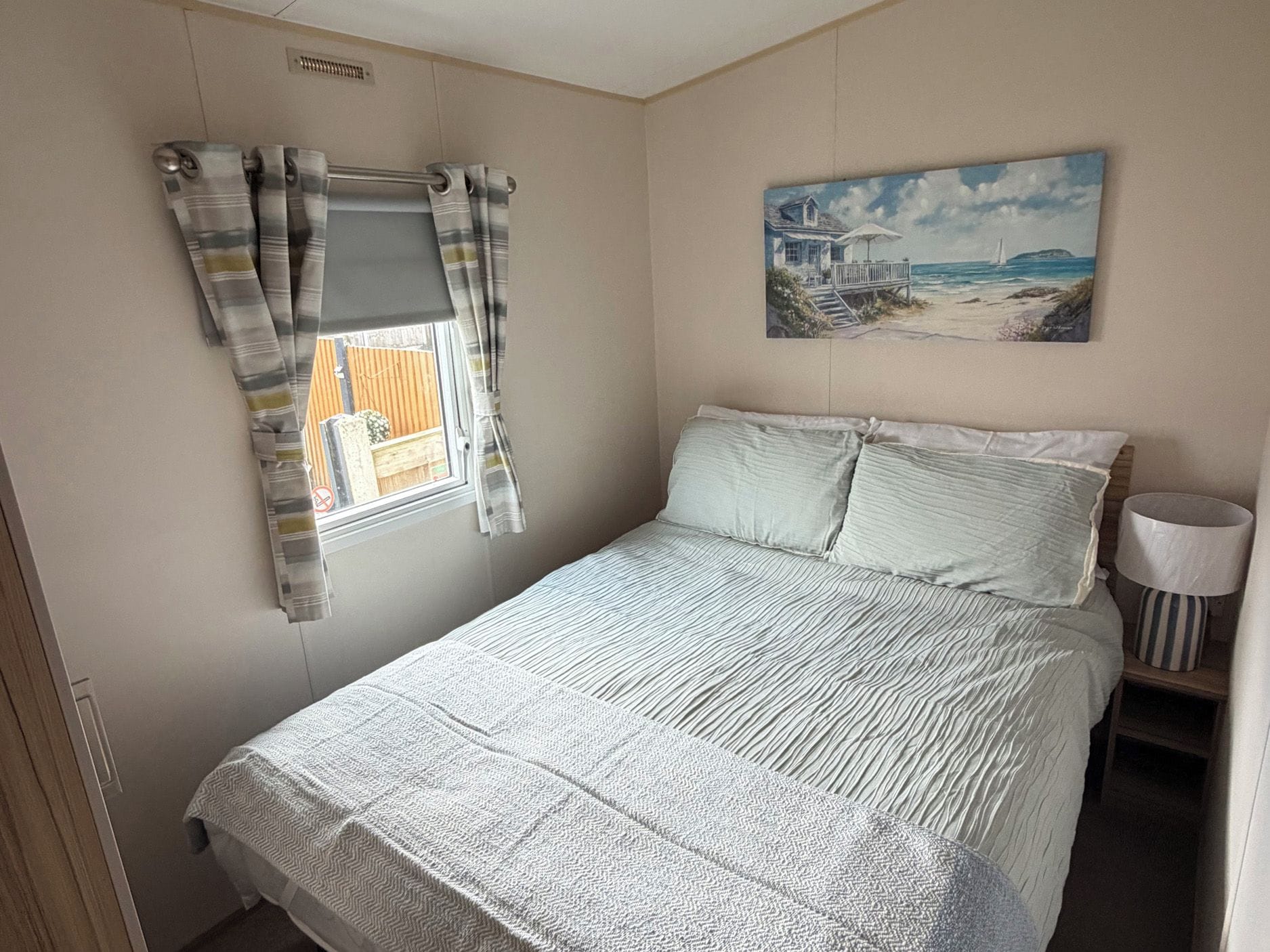 Quarto com cama, janela com cortinas, quadro na parede e luminária em uma mesinha de cabeceira.
