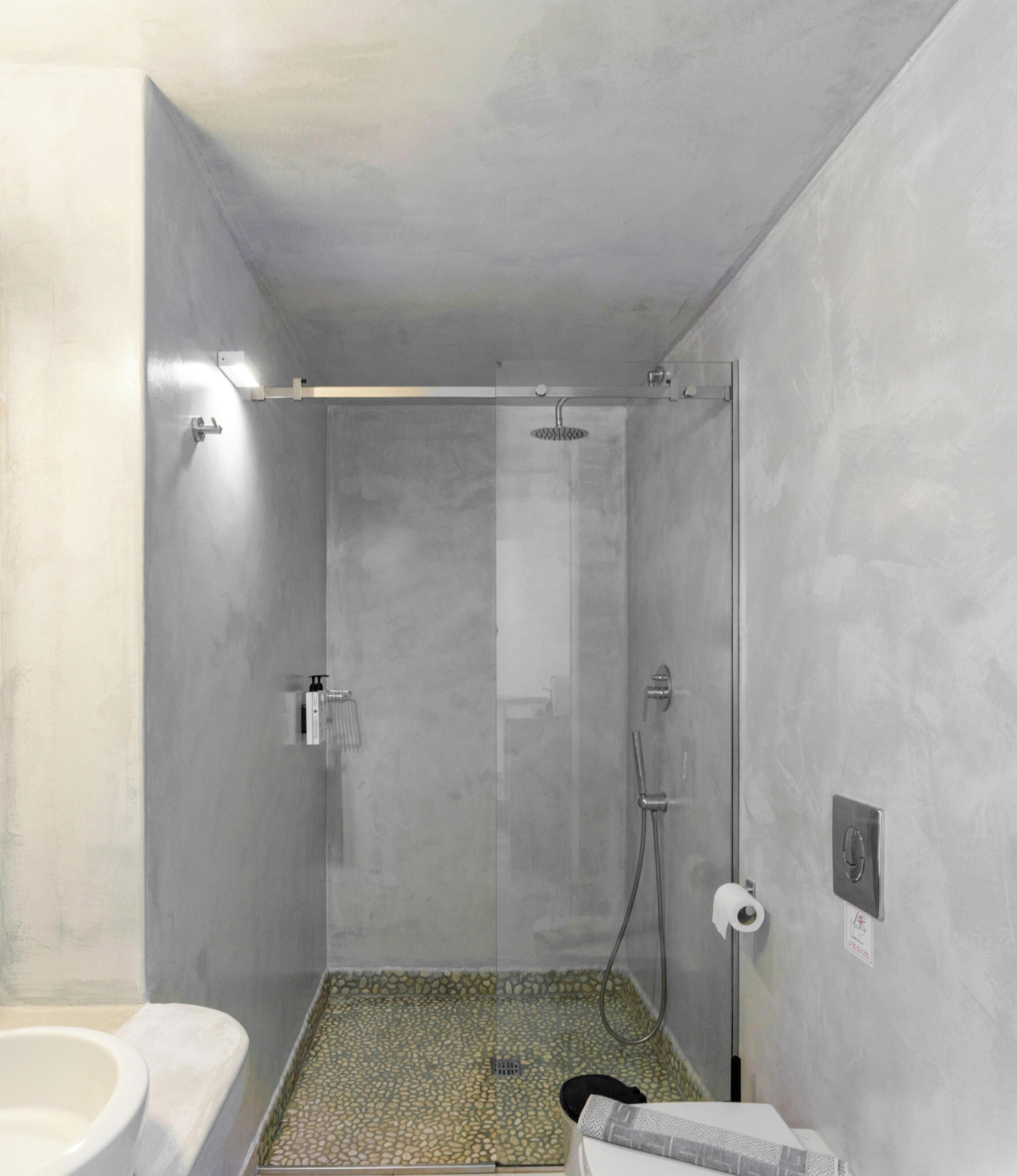 Chuveiro com porta de vidro, chuveiro manual, torneira fixada na parede e vaso sanitário à esquerda.