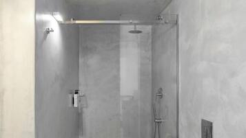 Chuveiro com porta de vidro, chuveiro manual, torneira fixada na parede e vaso sanitário à esquerda.