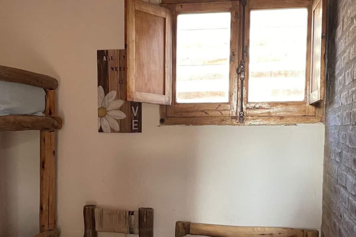 Quarto rústico com beliche de madeira, uma cadeira de madeira simples, uma cama pequena com travesseiro e uma janela com moldura de madeira.