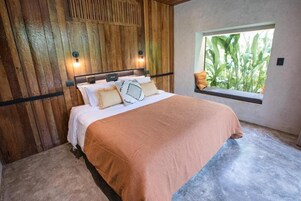 Quarto com parede de madeira, cama grande com manta rosa e janela com vista para vegetação.