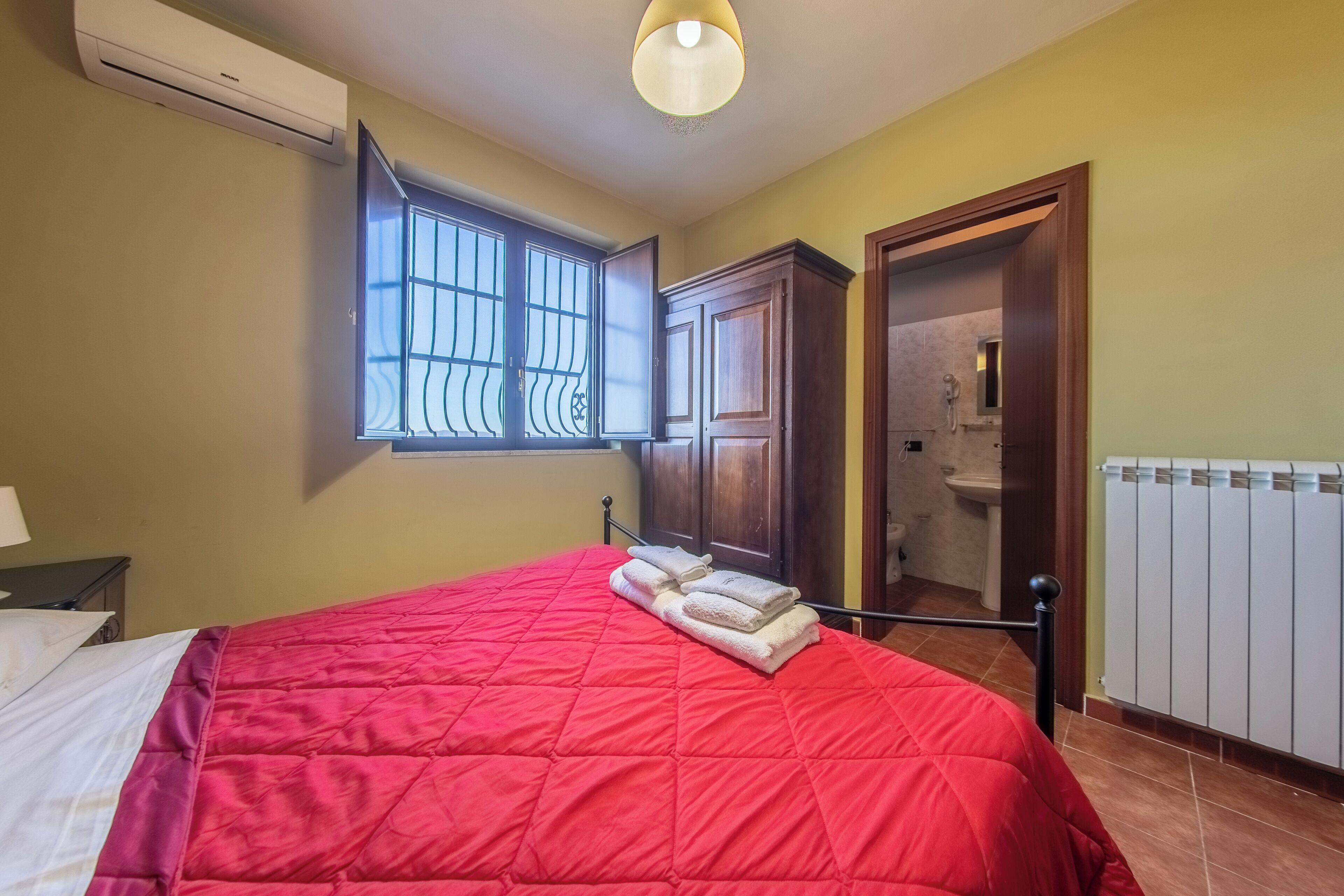 Quarto com colcha vermelha, janela com grades, guarda-roupa e banheiro visível através de uma porta aberta.