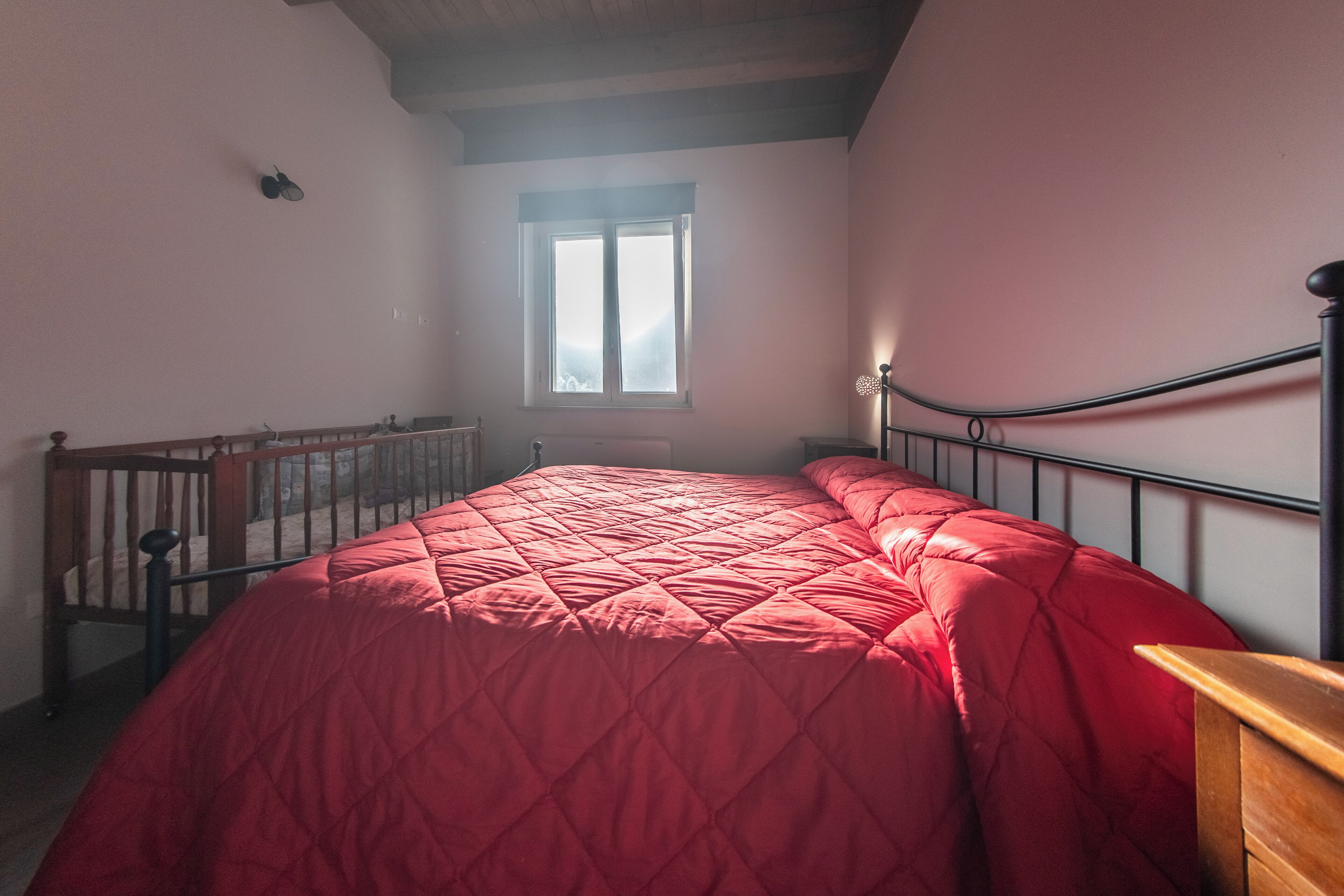 Quarto com colcha acolchoada vermelha, berço de madeira, janela com cortinas e vigas de madeira no teto.