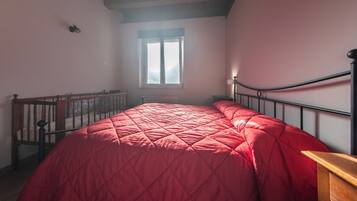 Quarto com colcha acolchoada vermelha, berço de madeira, janela com cortinas e vigas de madeira no teto.