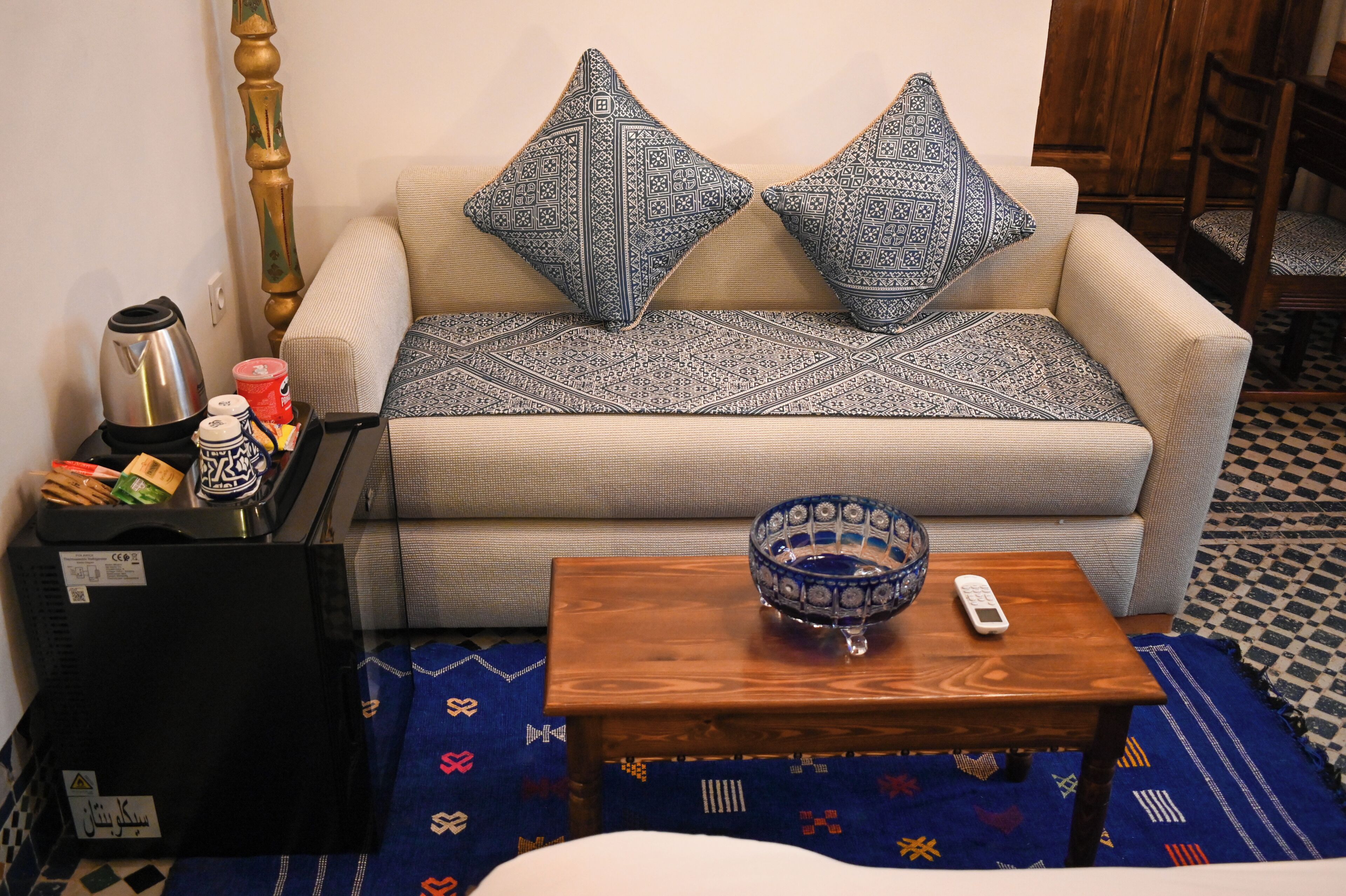 Uma sala de estar com sofá, almofadas estampadas, mesa de centro com uma tigela e controle remoto, tapete estampado e uma mesinha lateral com chaleira e petiscos.