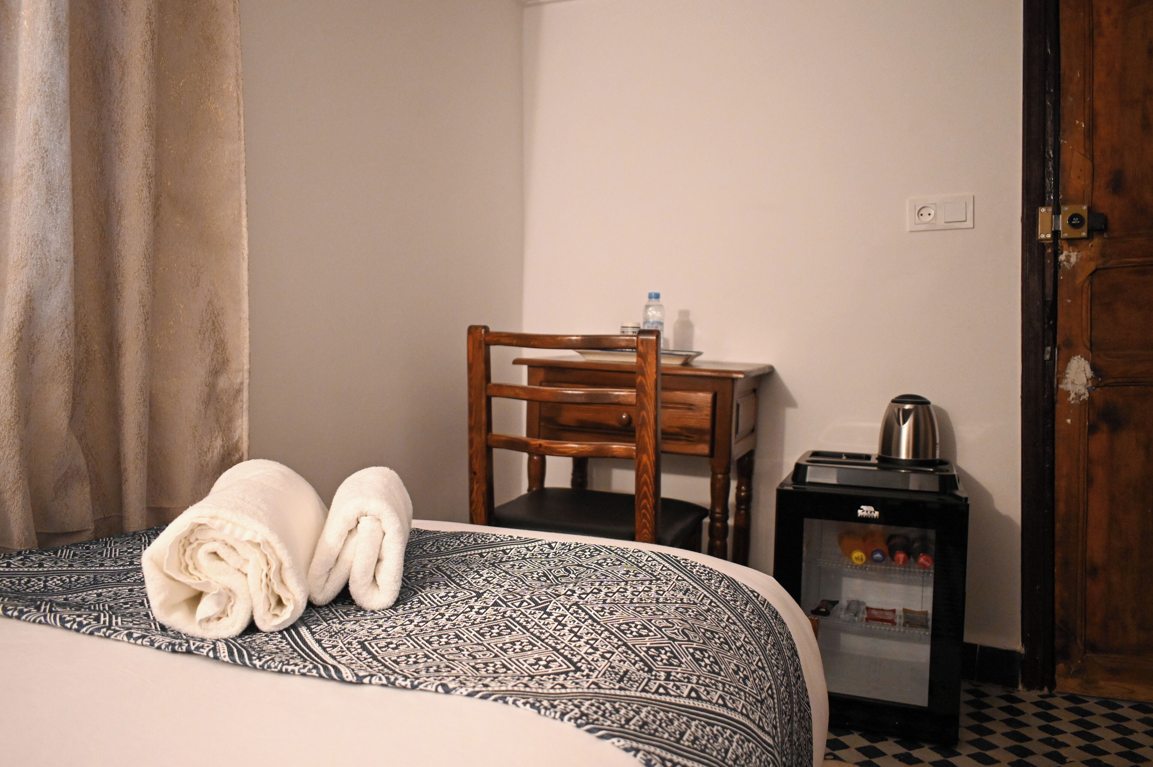 Quarto de hotel com uma cama, uma cadeira, uma mesinha com uma garrafa e uma chaleira, e um minibar.