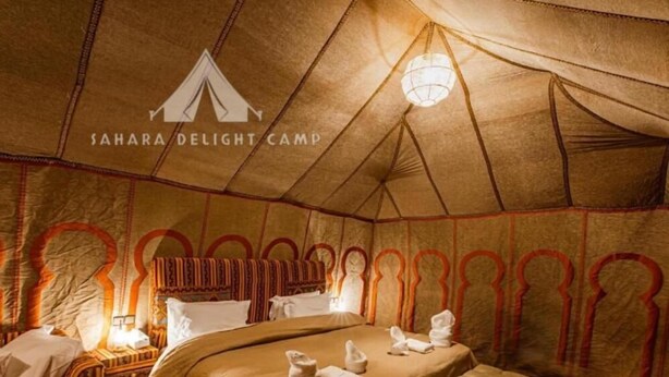Quarto coberto por tenda, com cama, banco e mesas de cabeceira, apresentando um papel de parede estampado e um candelabro.
