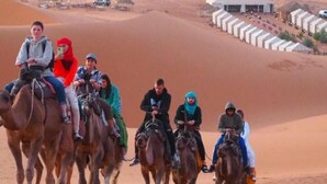 Um grupo de pessoas a cavalgar camelos em paisagem desértica, com tendas e um edifício ao fundo.