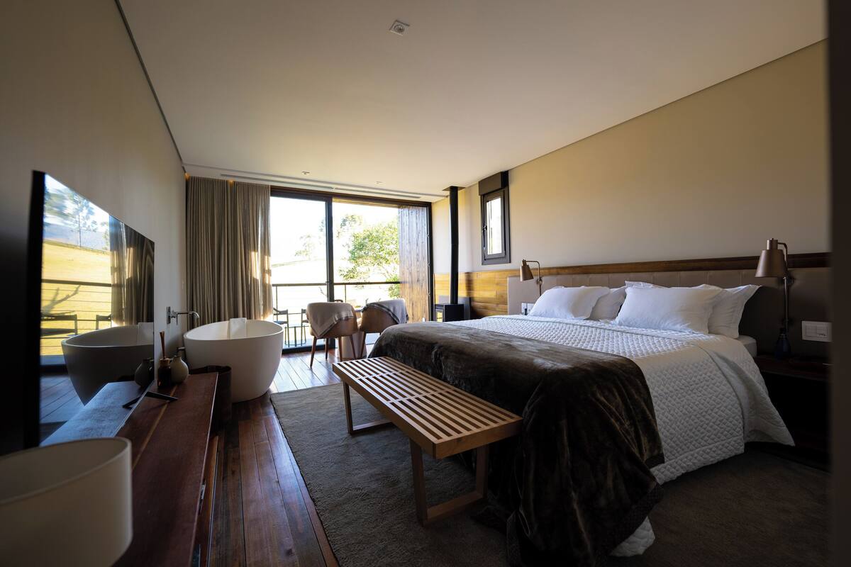 Quarto de hotel moderno com uma cama grande, um banco de madeira, uma mesa com abajur e uma janela ampla com vista para o exterior.