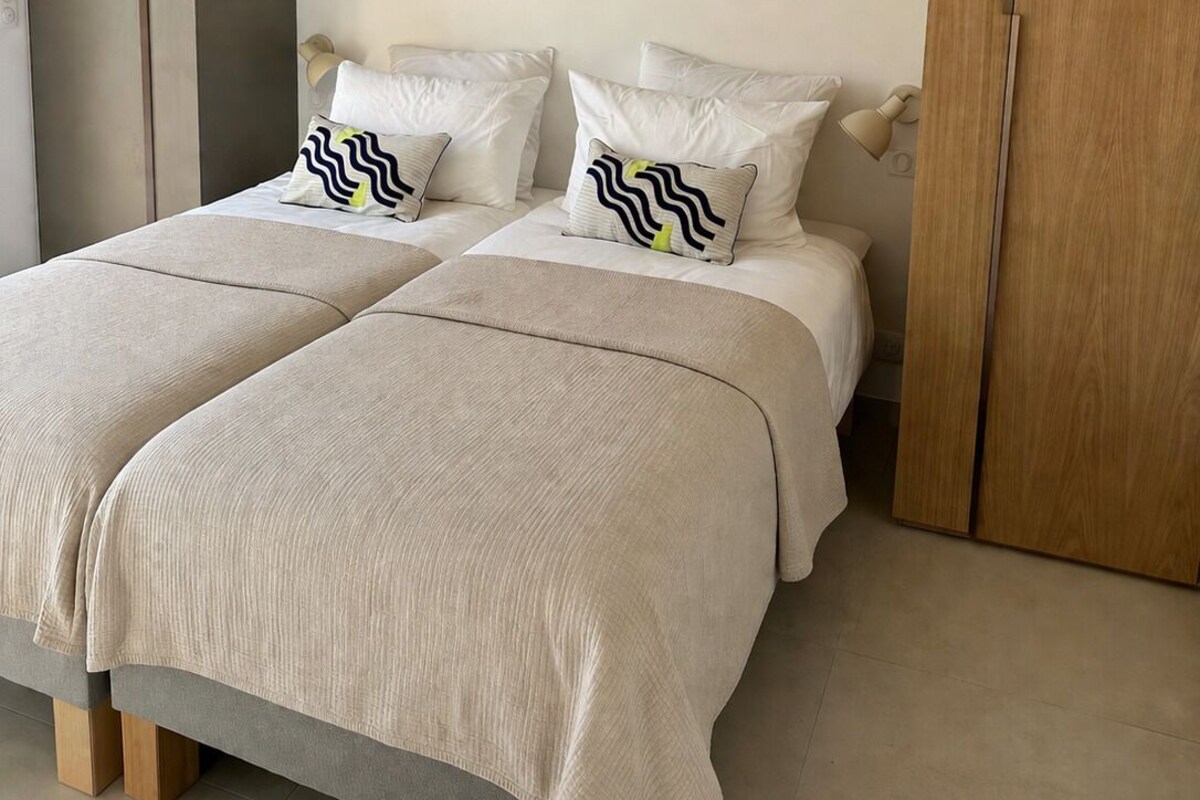 Uma cama de casal com duas almofadas, cabeceira cinzenta e guarda-roupa de madeira.