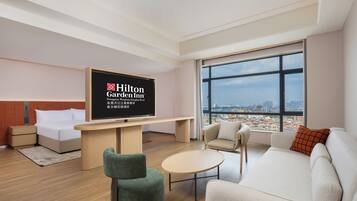 Quarto de hotel moderno com cama, escrivaninha, televisão, sofá e uma grande janela com vista para a cidade.