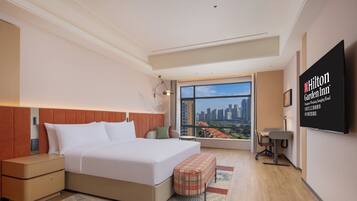Quarto de hotel moderno com uma cama grande, uma escrivaninha, uma cadeira, uma televisão e uma janela com vista para a cidade.