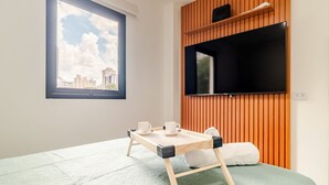 Quarto de hotel com cama, janela com vista para a cidade, televisão de tela plana fixada em um painel de madeira e mesinha de bandeja de madeira com xícaras e pires.