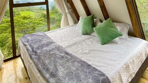 Quarto amplo com pé direito alto, uma cama grande, vigas de madeira e vista para o verde lá fora.