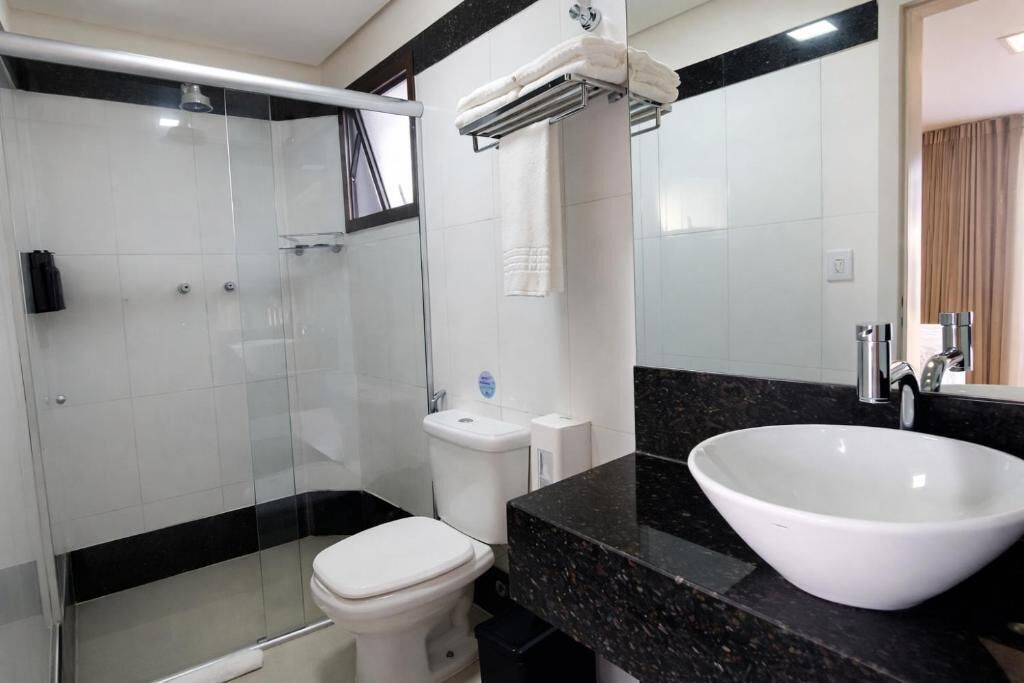 Uma casa de banho moderna com cabine de duche em vidro, sanita branca, lavatório branco e bancada preta.