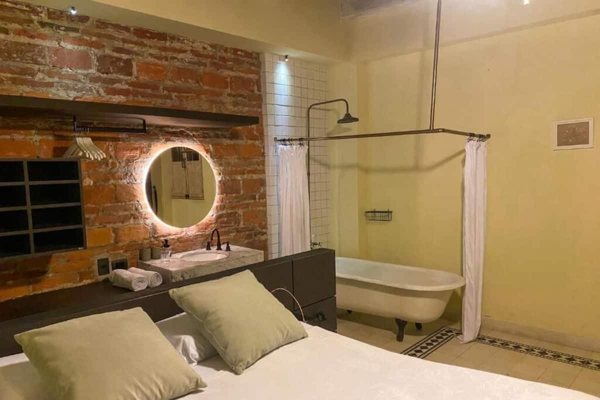 Quarto com parede de tijolos, espelho redondo, cama com travesseiros, mesa de cabeceira, banheiro com banheira, chuveiro e porta-toalhas.