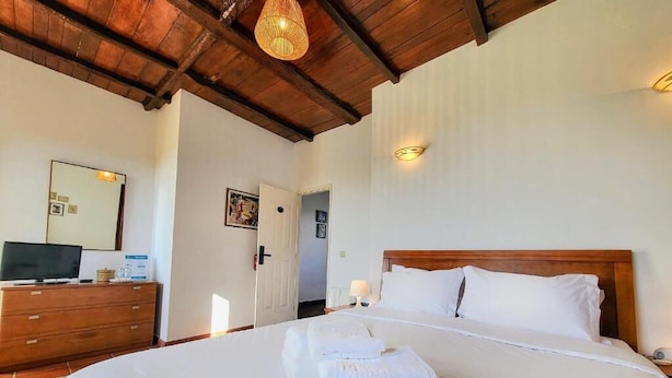 Quarto de hotel com uma cama grande, cabeceira de madeira, roupa de cama branca, cômoda de madeira, televisão e espelho.