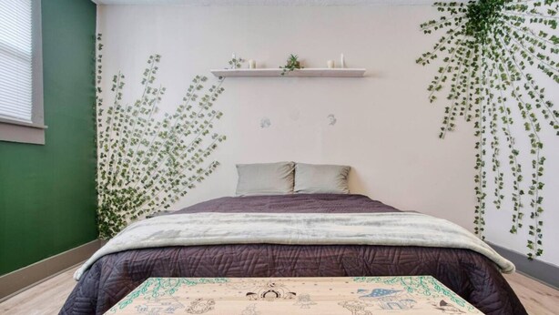 Quarto com cama, mesa de cabeceira com detalhe decorativo e plantas pendentes verdes nas paredes.