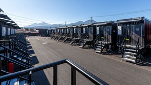 Fileira de cabanas compactas e modernas, com guarda-corpos de metal e portas numeradas, com montanhas e céu claro ao fundo.