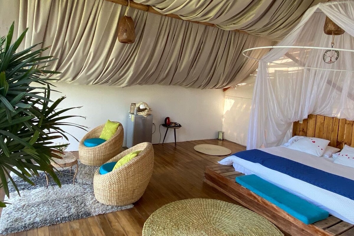 Um quarto com uma cama, duas cadeiras de vime, uma mesinha e uma planta grande.