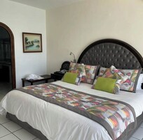 Uma cama bem arrumada com um edredom estampado e várias almofadas, uma cabeceira escura, uma mesa de cabeceira com abajur e um quadro na parede.