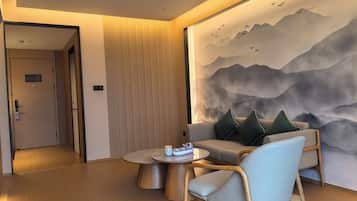 Quarto de hotel moderno com piso de madeira, um sofá com almofadas verdes, uma mesa redonda com chaleira e xícaras, e um mural grande na parede com montanhas e pássaros.