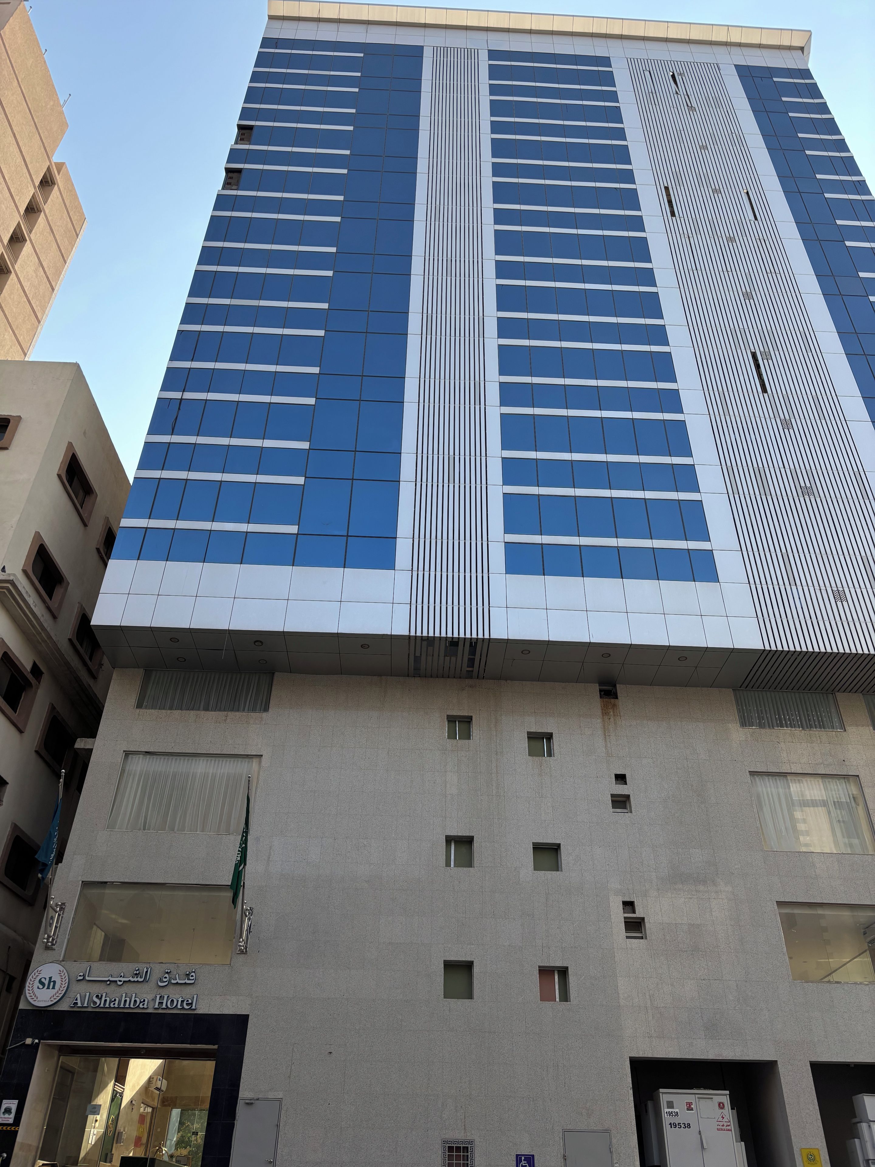 Um edifício alto com design arquitetônico moderno, destacando uma faixa vertical de janelas e uma entrada no nível da rua com a placa do Hotel Al Shahba.