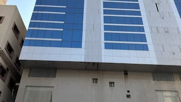 Um edifício alto com design arquitetônico moderno, destacando uma faixa vertical de janelas e uma entrada no nível da rua com a placa do Hotel Al Shahba.